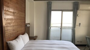 Quarto de hotel com cabeceira de madeira, cama grande com roupa de cama branca, mesa de cabeceira pequena, luminária fixada na parede, janela com cortinas e vista para uma varanda.