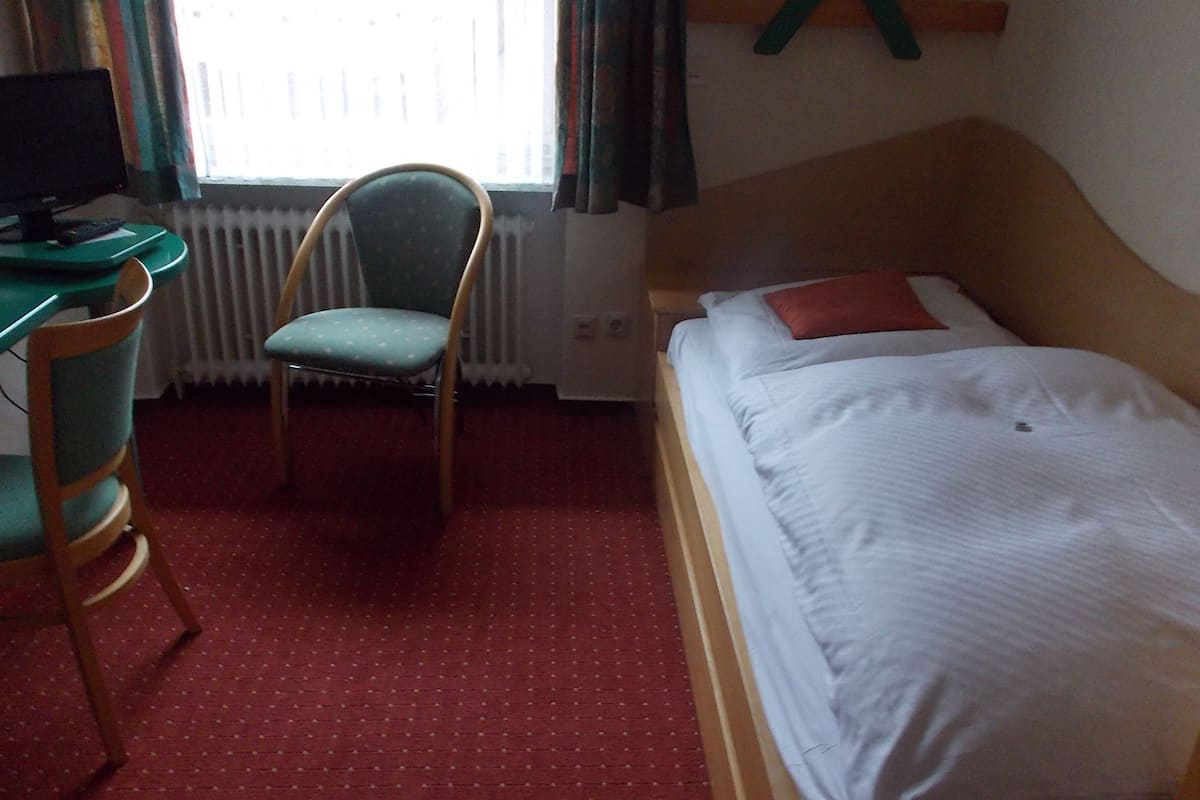 Quarto de hotel com uma cama de solteiro, uma escrivaninha com computador, uma cadeira e um radiador.