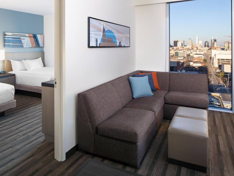 Quarto de hotel moderno com uma janela grande que oferece vista para a cidade, um sofá modular, uma cama e uma pequena mesa.