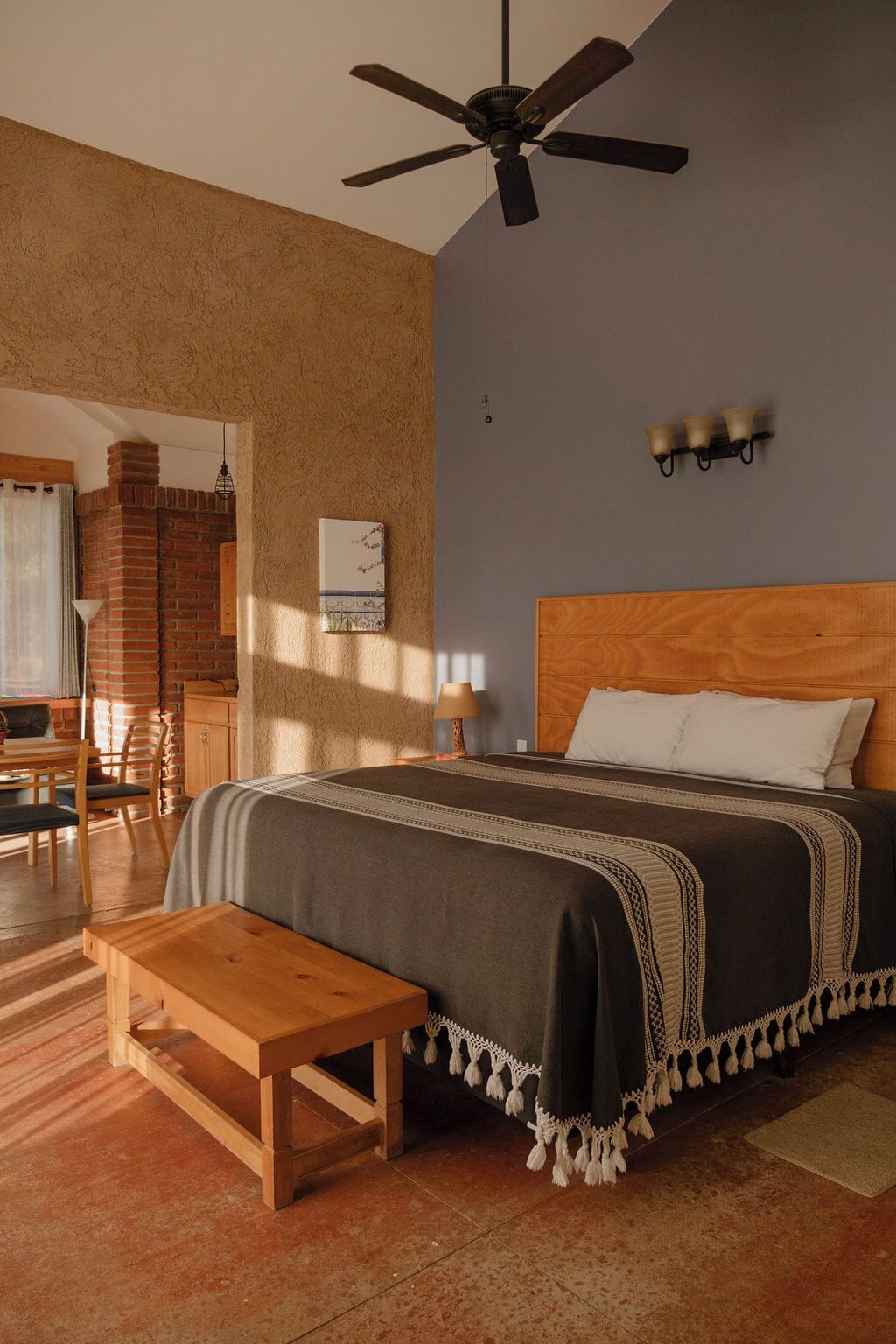Quarto com cama de madeira, um banquinho, ventoinha no teto e parede de tijolos.