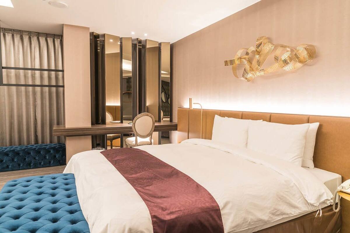 Quarto de hotel com cama grande, escrivaninha, cadeira e um banquinho azul com capitonê.