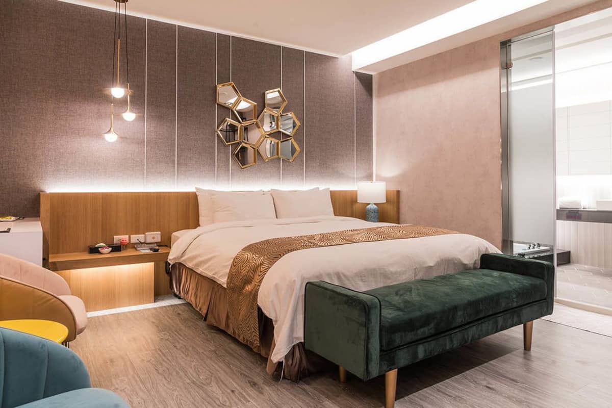 Quarto de hotel moderno com uma cama grande, mesas de cabeceira, um banco verde e um espelho fixado na parede.
