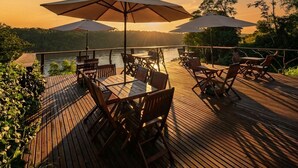 Um deck de madeira com mesas, cadeiras, guarda-sóis e vista para a água e colinas.