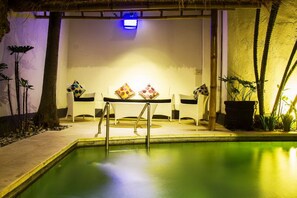 Área de piscina com mesa e cadeiras, cercada por plantas e uma estrutura com telhado de palha.