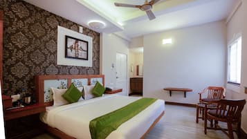 Quarto com cama, mesa de cabeceira, cadeira, ventoinha no teto e banheiro visível por uma porta aberta.