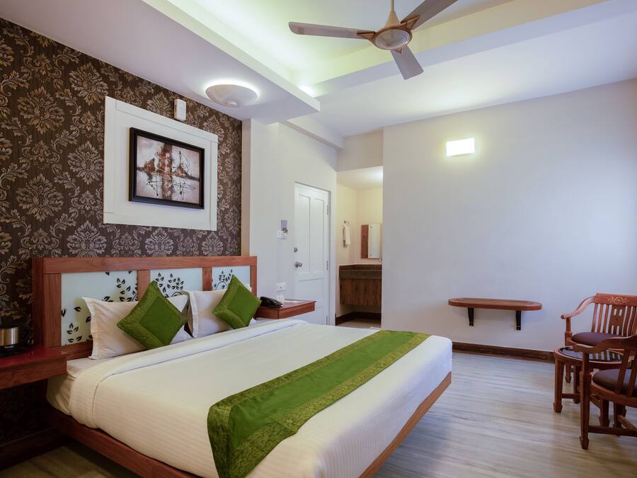 Quarto com cama, mesa de cabeceira, cadeira, ventoinha no teto e banheiro visível por uma porta aberta.