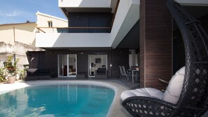 Uma área moderna com piscina ao ar livre, uma espreguiçadeira, uma mesa de jantar e uma cozinha visível através das portas de vidro.