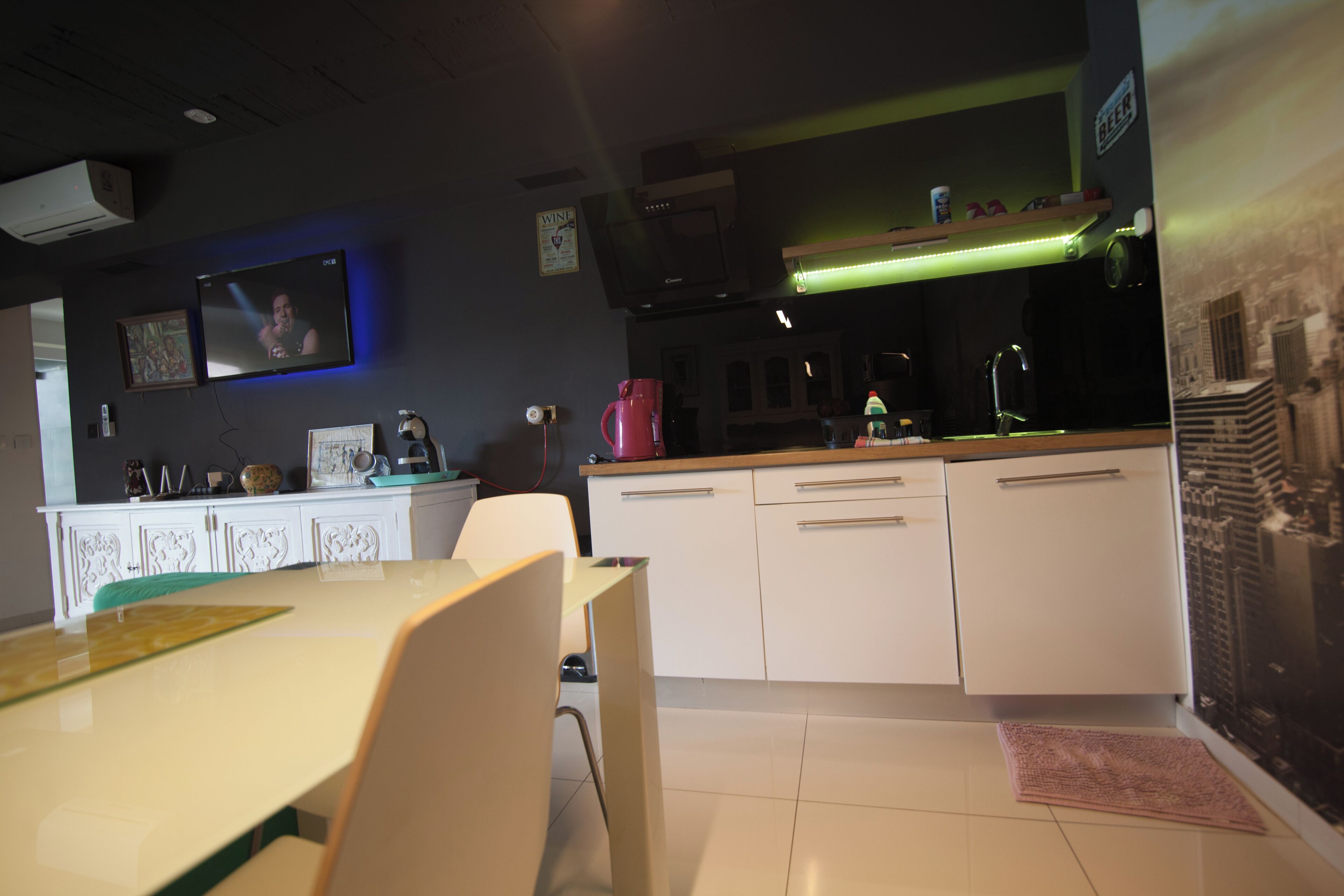 Uma cozinha moderna com armários brancos, uma área de jantar com mesa e cadeiras, uma televisão fixada na parede e um mural com vista para a cidade.