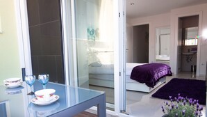 Quarto de hotel moderno com porta de vidro que dá para uma varanda, cama com manta roxa, banheiro pequeno e área de jantar com mesa e cadeiras.