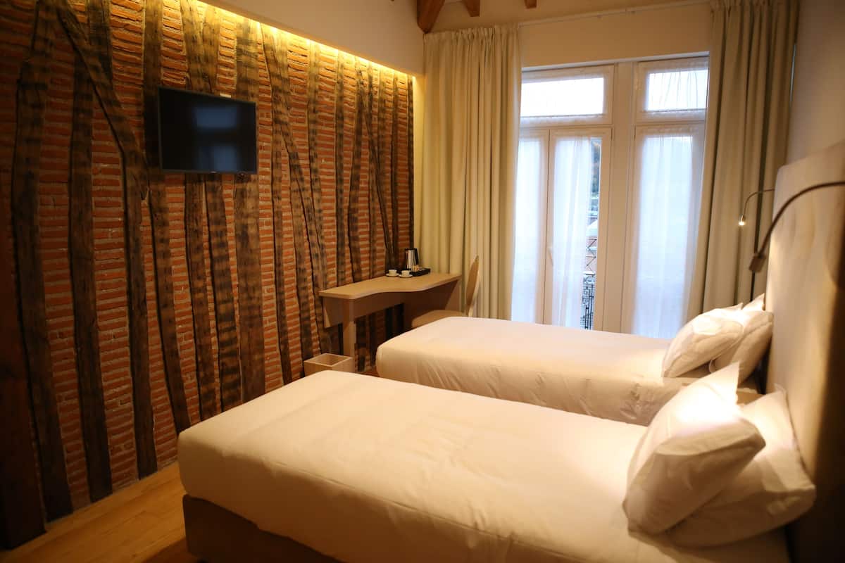 Quarto de hotel com duas camas, uma televisão de tela plana presa a uma parede com textura de tijolos, uma pequena mesa de trabalho com cadeira e uma janela com cortinas.