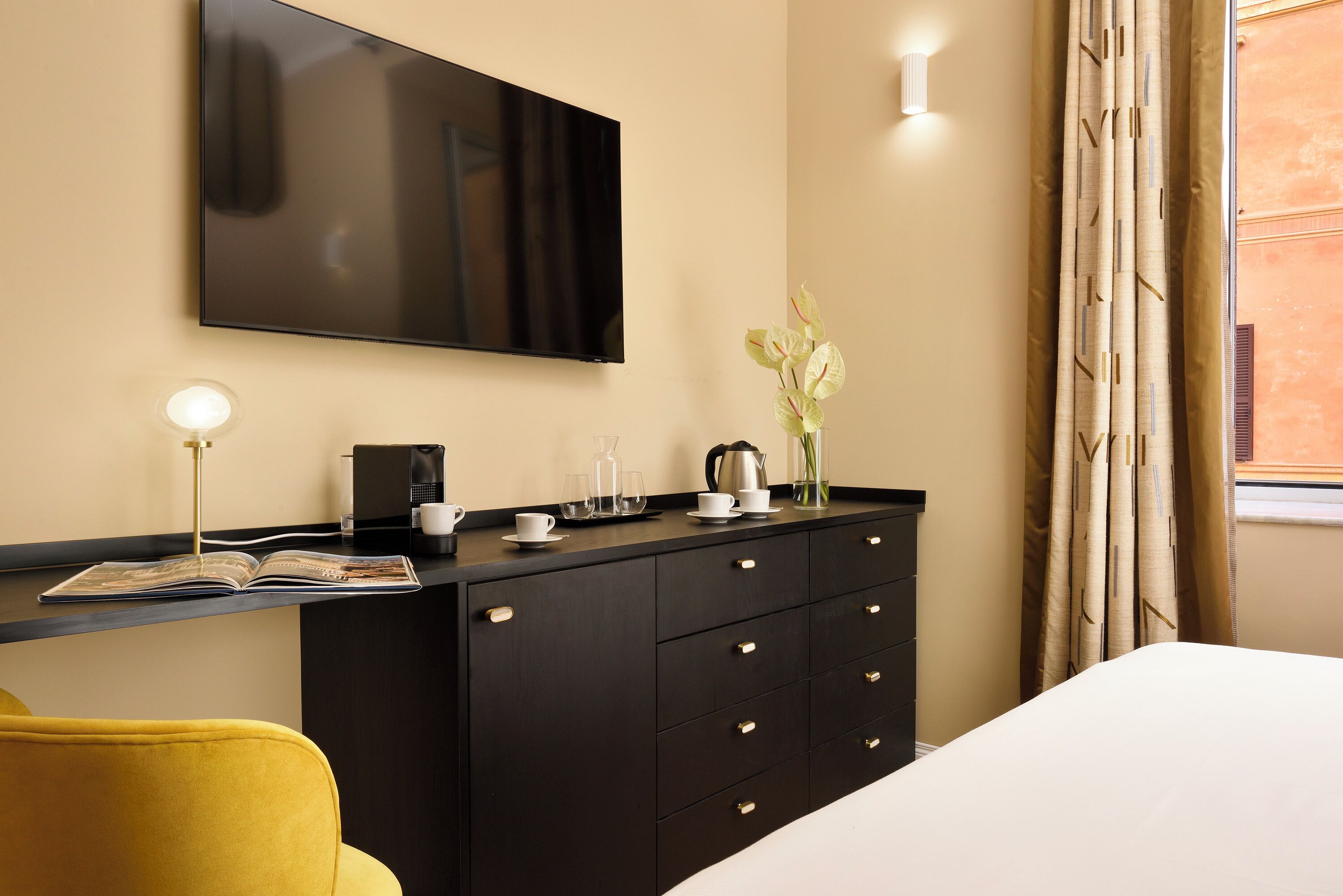 Quarto de hotel com televisão de tela plana na parede, cômoda de madeira escura com gavetas, poltrona amarela e janela com cortinas.