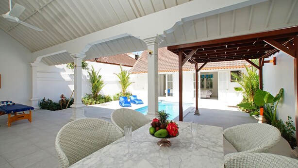 Uma área de estar coberta ao ar livre, com mesa posta para dois, rodeada por cadeiras de vime, uma piscina e um edifício branco ao fundo.