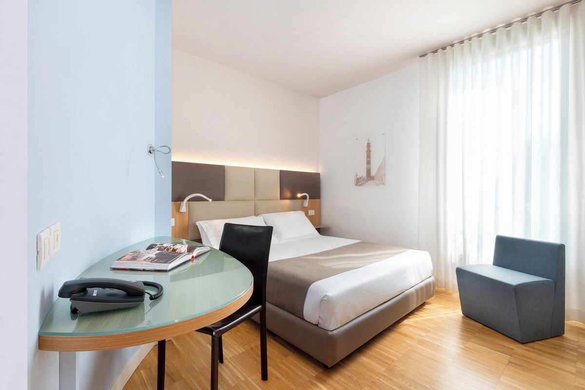 Quarto de hotel com cama, escrivaninha, telefone, cadeira e janela com cortinas.