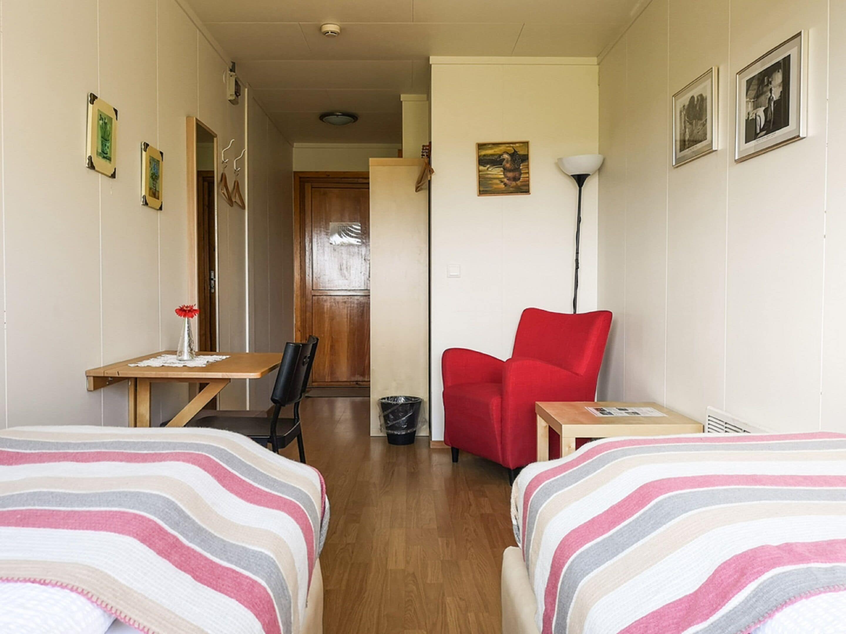 Quarto de hotel com duas camas, uma escrivaninha, uma cadeira, uma luminária e uma porta que dá para outro cômodo.