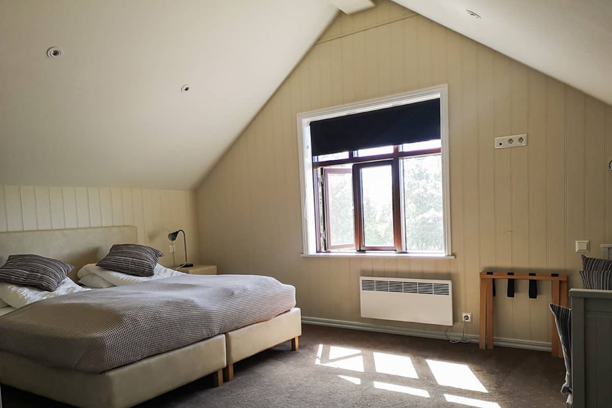 Quarto com teto inclinado, uma janela grande, cama, mesa de cabeceira e radiador.