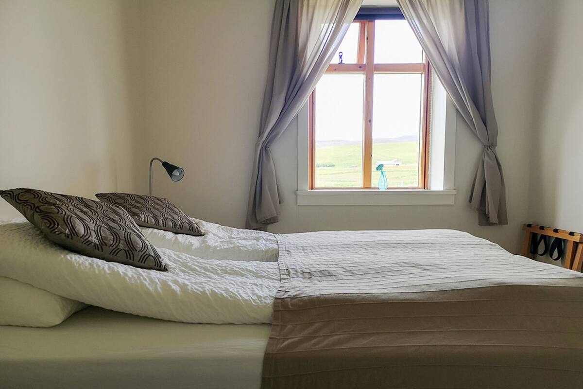 Uma cama com almofadas estampadas, uma luminária de cabeceira e vista para um campo através de uma janela com cortinas.