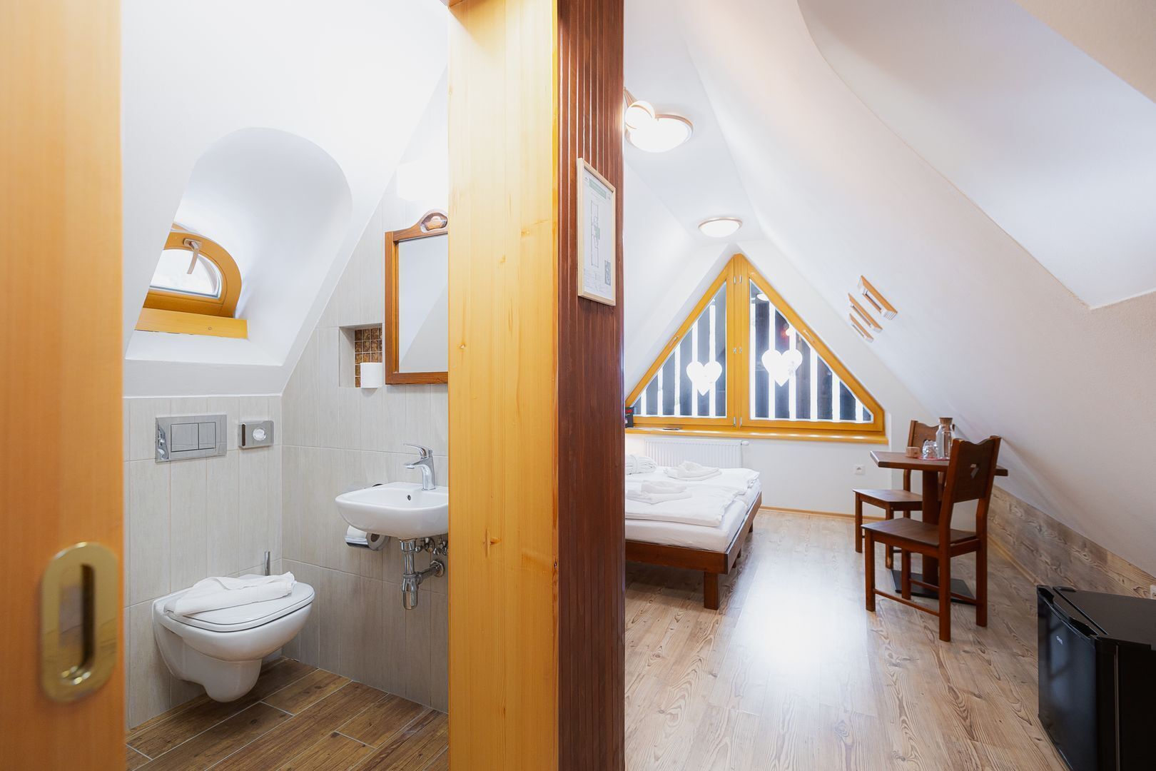 Quarto com teto inclinado, cama, janela com vitrais em formato de coração e piso de madeira.