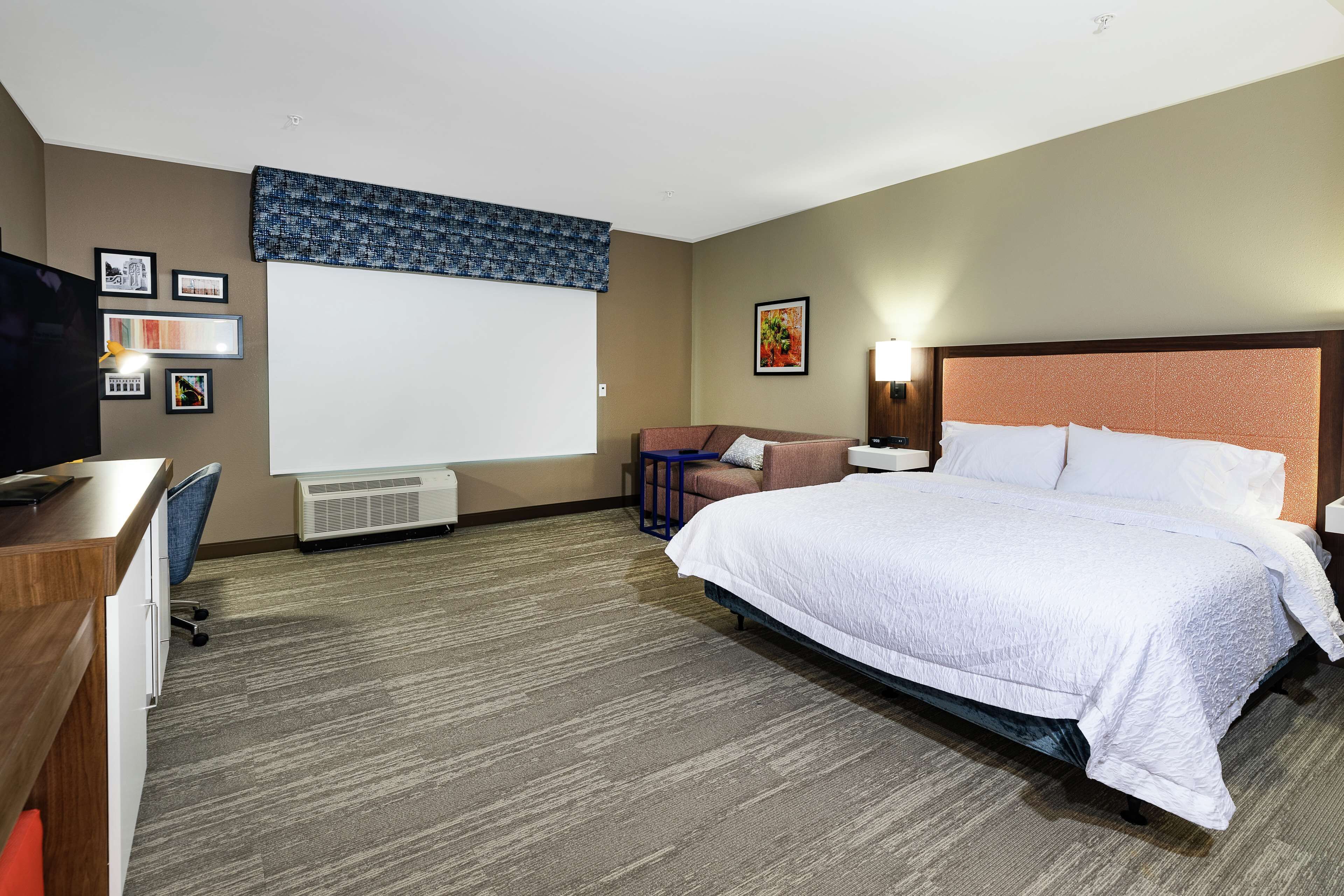 Quarto de hotel com uma cama grande, uma escrivaninha com um computador, uma cadeira, uma tela de projetor e um ar condicionado fixado na parede.