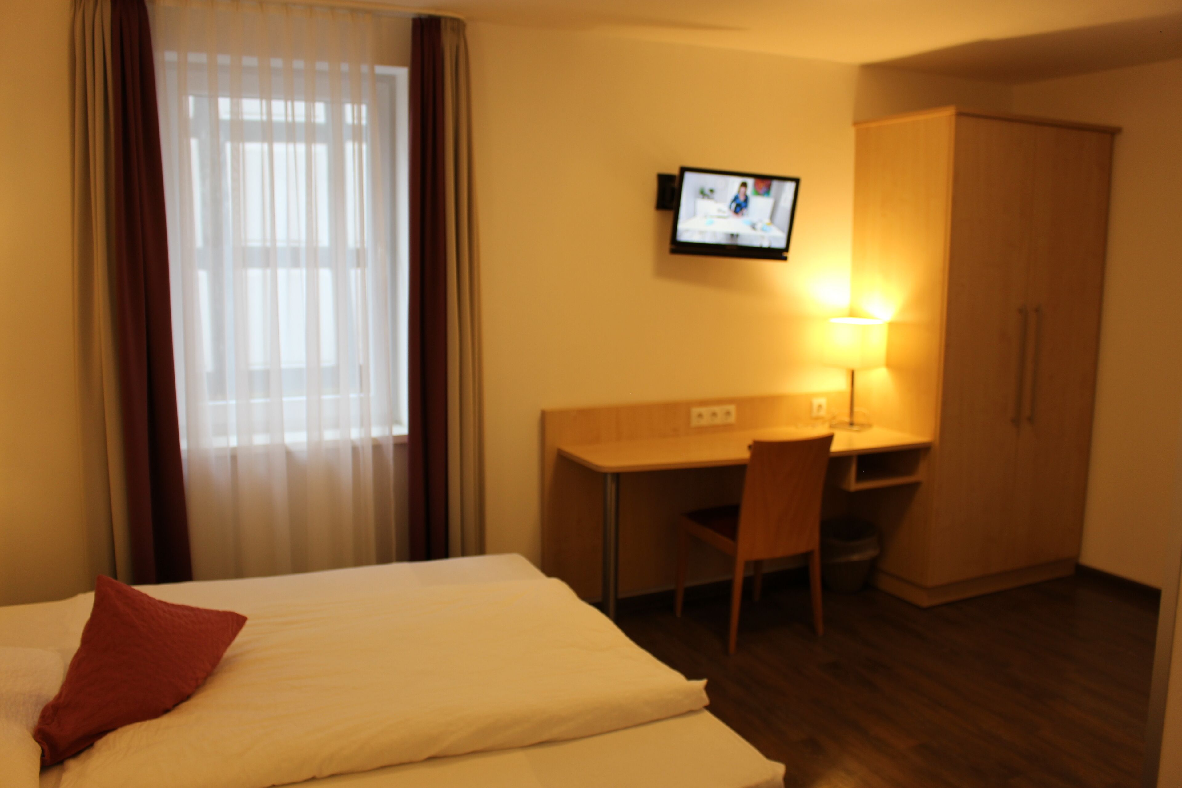 Quarto de hotel com cama, escrivaninha, cadeira, televisão e guarda-roupa.