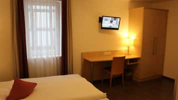 Quarto de hotel com cama, escrivaninha, cadeira, televisão e guarda-roupa.