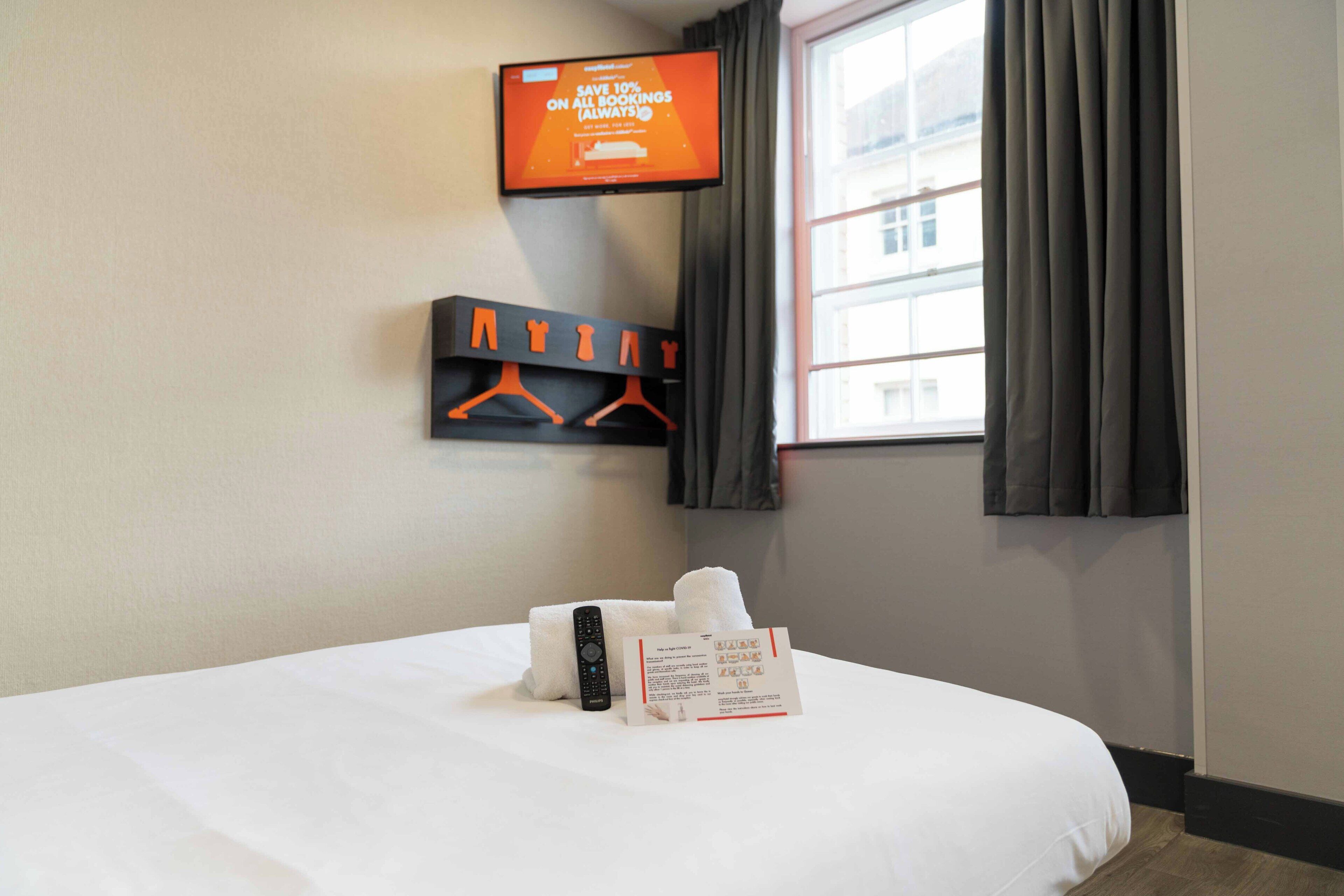 Quarto de hotel com cama, controle remoto, toalha, cartão de boas-vindas, televisão na parede e janela com cortinas.