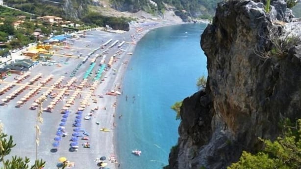 Uma praia costeira com fileiras de guarda-sóis e espreguiçadeiras, um penhasco rochoso e um mar azul cristalino.