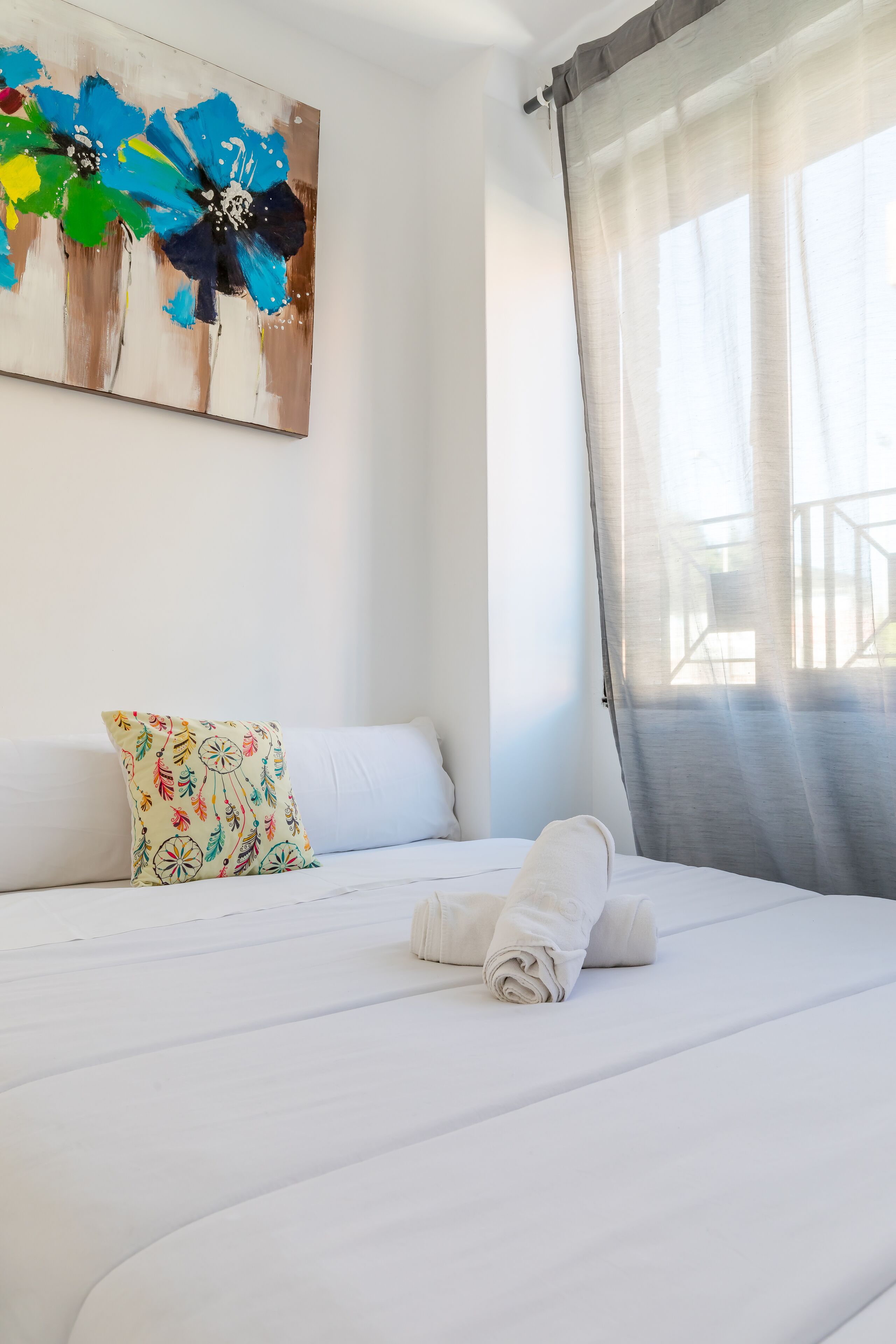 Cama bem arrumada com lençóis brancos, toalha enrolada, almofada colorida e um grande quadro abstrato na parede.