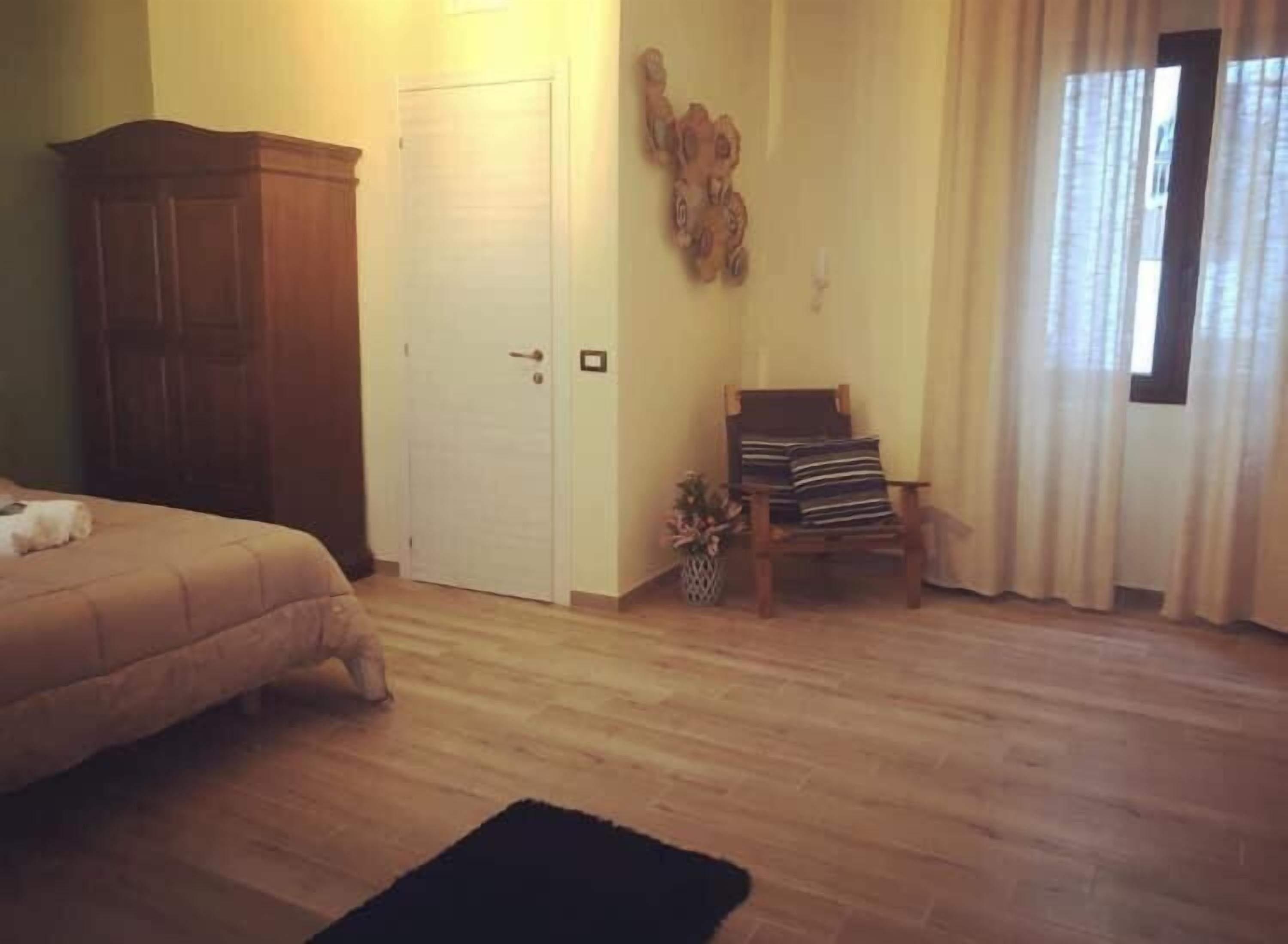 Quarto com cama, guarda-roupa, cadeira com almofadas, porta e janela com cortinas.