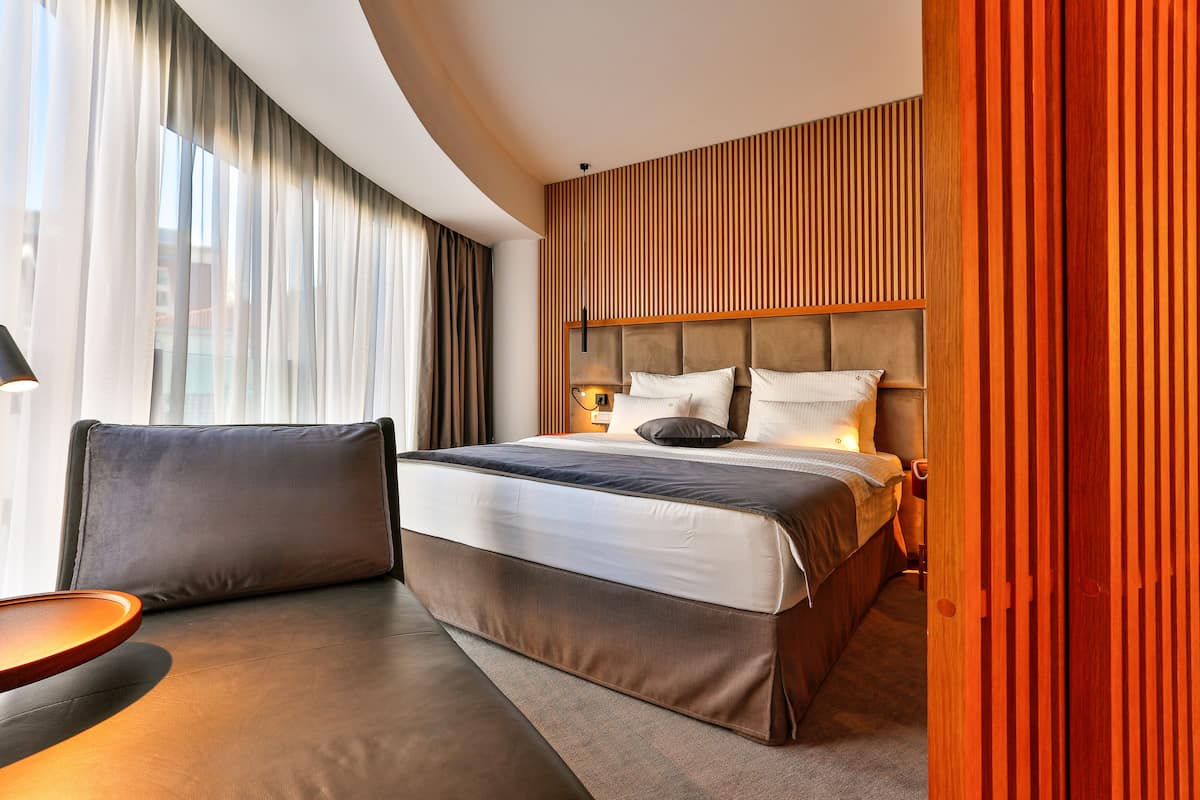 Quarto de hotel moderno com uma cama grande, cabeceira de madeira, área de estar com mesa e vista da cidade por grandes janelas.