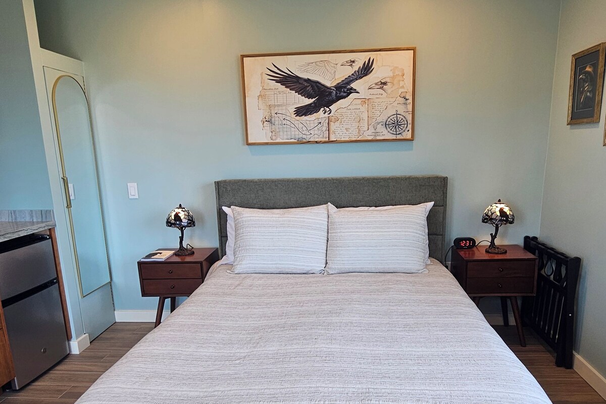 Quarto com uma cama grande, duas mesas de cabeceira com abajures, uma pintura de um pássaro na parede e uma porta à esquerda.