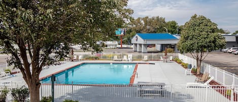 Piscina cercada por um muro branco, cadeiras de descanso e um edifício com telhado azul ao fundo.
