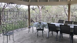 Um alpendre coberto com uma mesa e cadeiras, cerca de metal e árvores ao fundo.