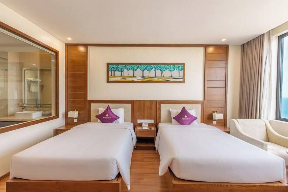 Quarto de hotel com duas camas, um quadro na parede, uma janela e uma cadeira.