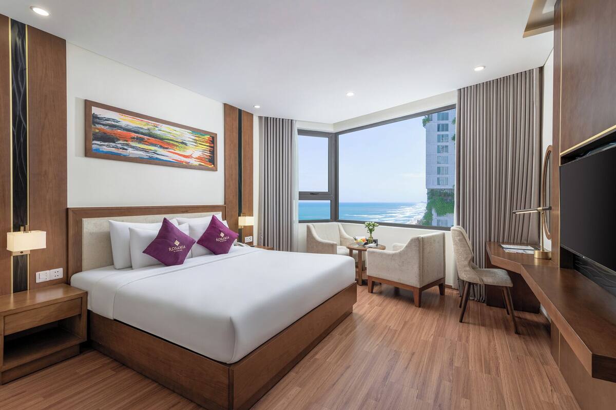 Quarto de hotel com cama grande, escrivaninha, cadeira, televisão e vista para o oceano.