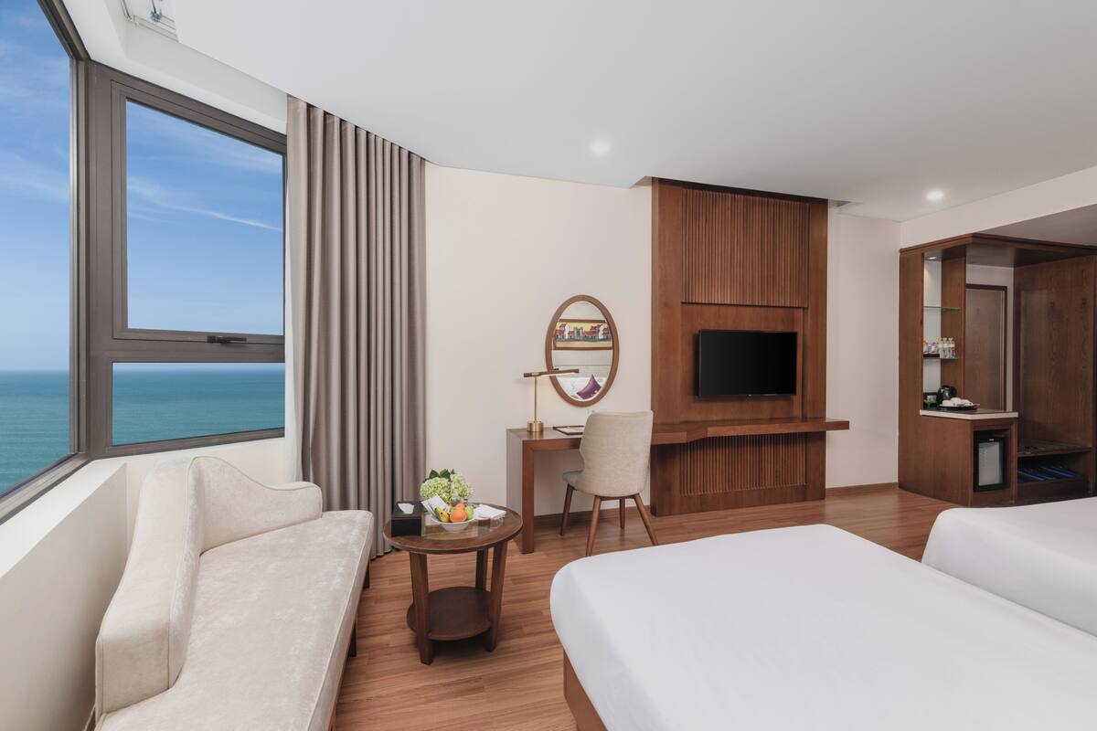 Quarto de hotel com uma janela grande, um sofá, uma escrivaninha com cadeira, uma televisão e um guarda-roupa.