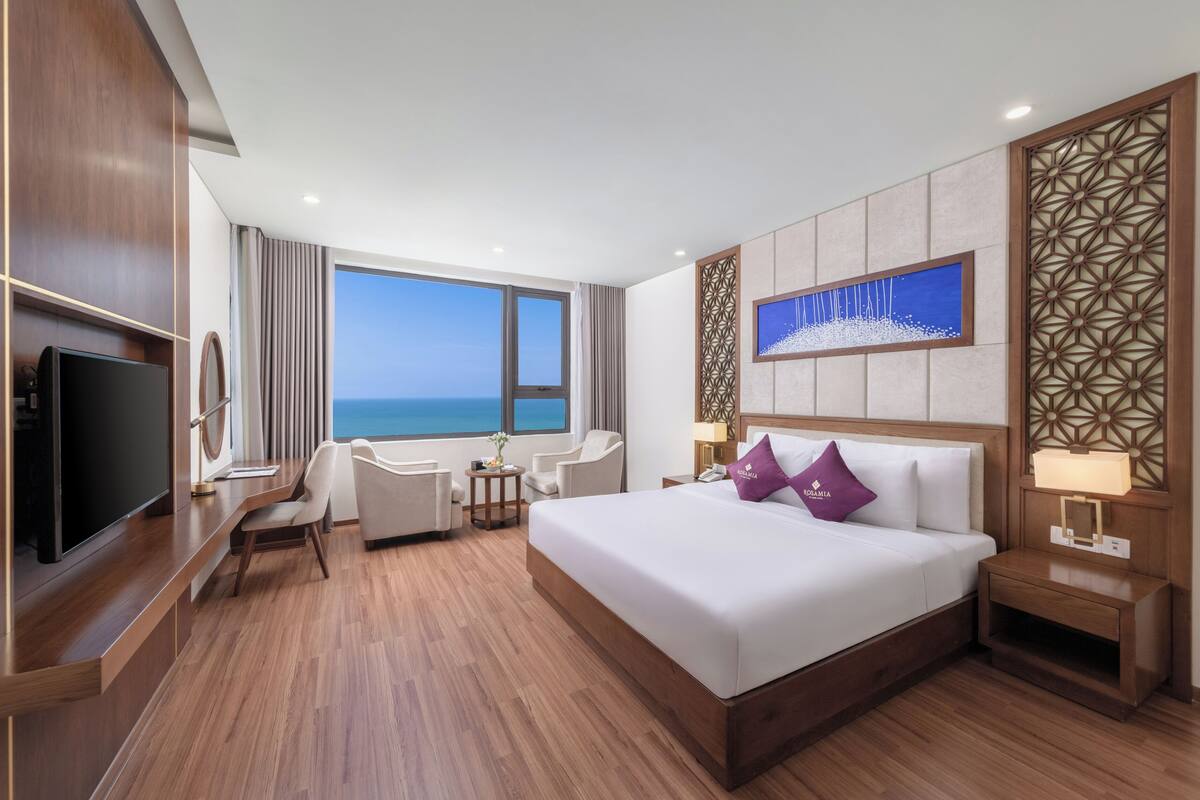Quarto de hotel com uma cama grande, televisão, escrivaninha e vista para o oceano.
