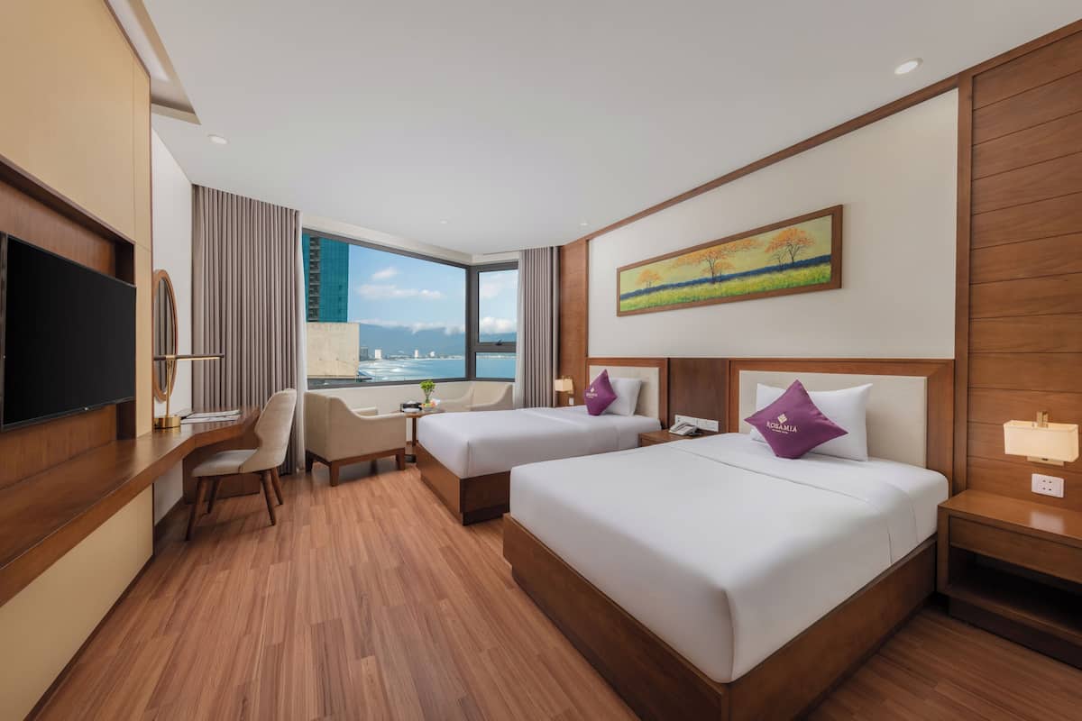 Quarto de hotel com duas camas, uma mesa, uma cadeira, televisão e uma grande janela com vista para a cidade.
