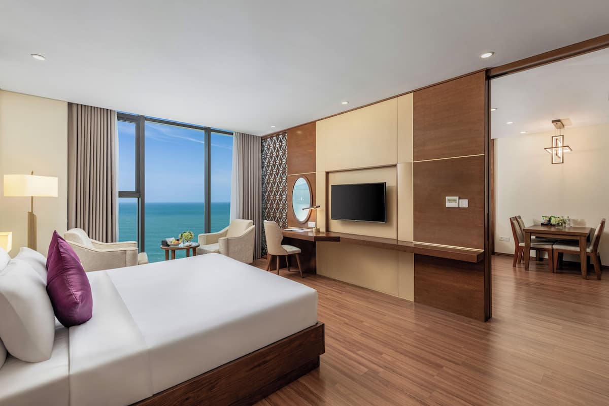 Quarto de hotel moderno com uma cama grande, escrivaninha, televisão e área de refeições com vista para o oceano.