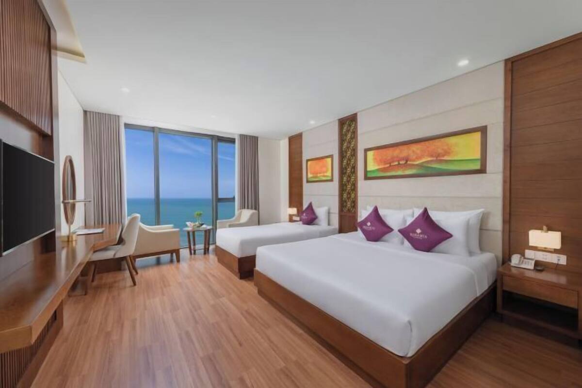 Quarto de hotel com duas camas, televisão, escrivaninha e vista para o mar.