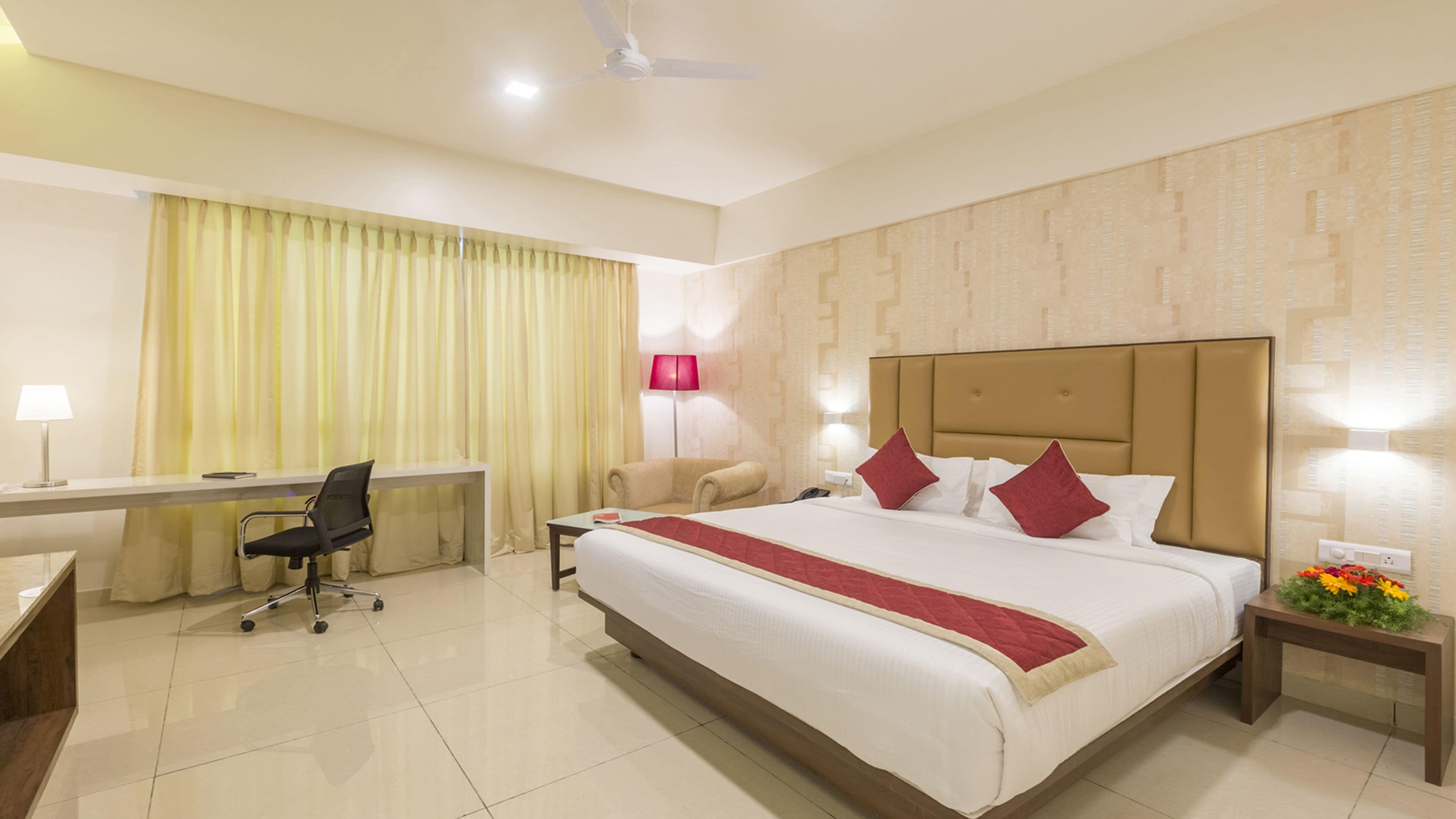 Quarto de hotel com uma cama grande, uma mesa, uma cadeira e uma mesinha de cabeceira com flores.