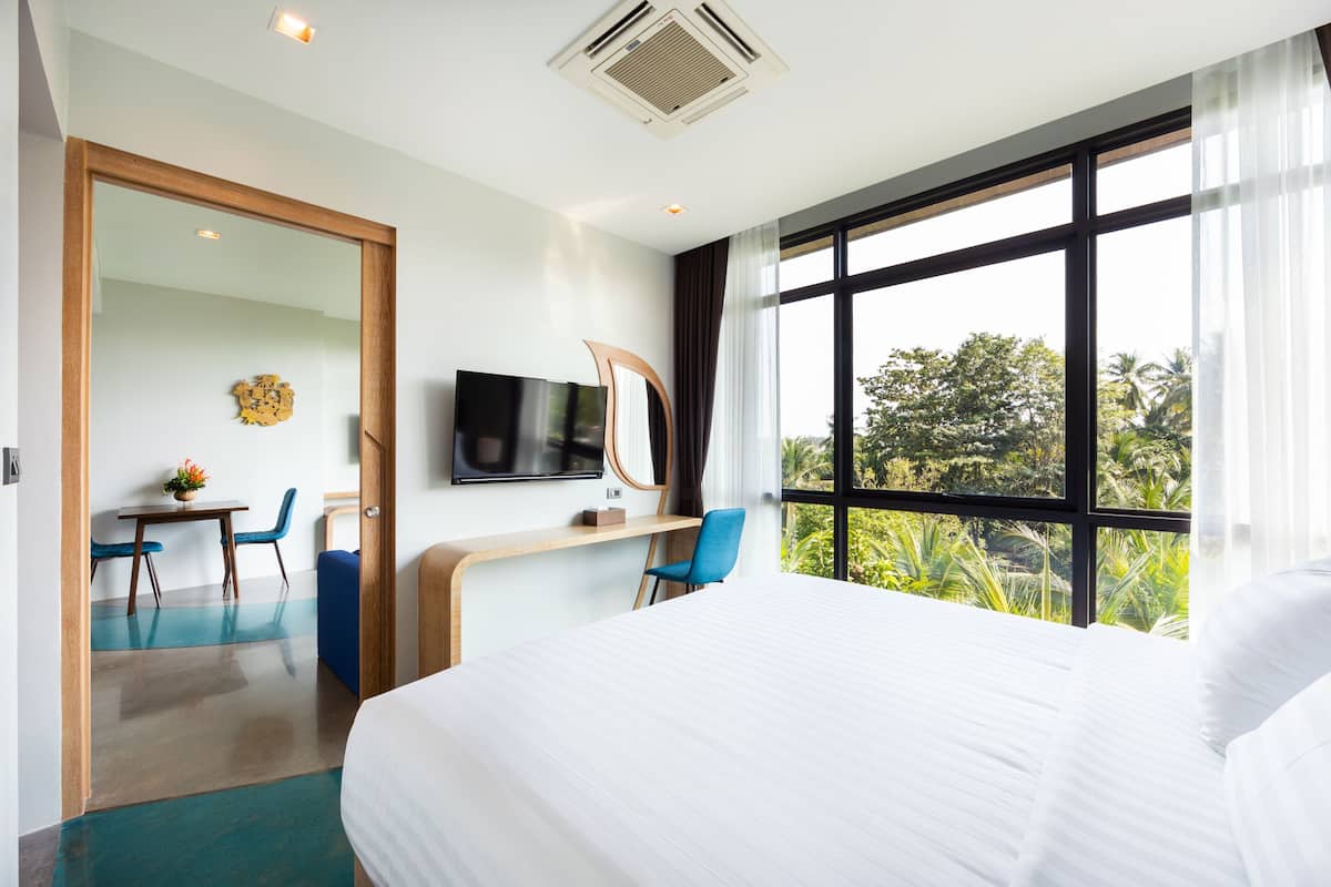 Quarto de hotel com uma cama grande, televisão na parede, escrivaninha com cadeira e vista para o verde pela janela.
