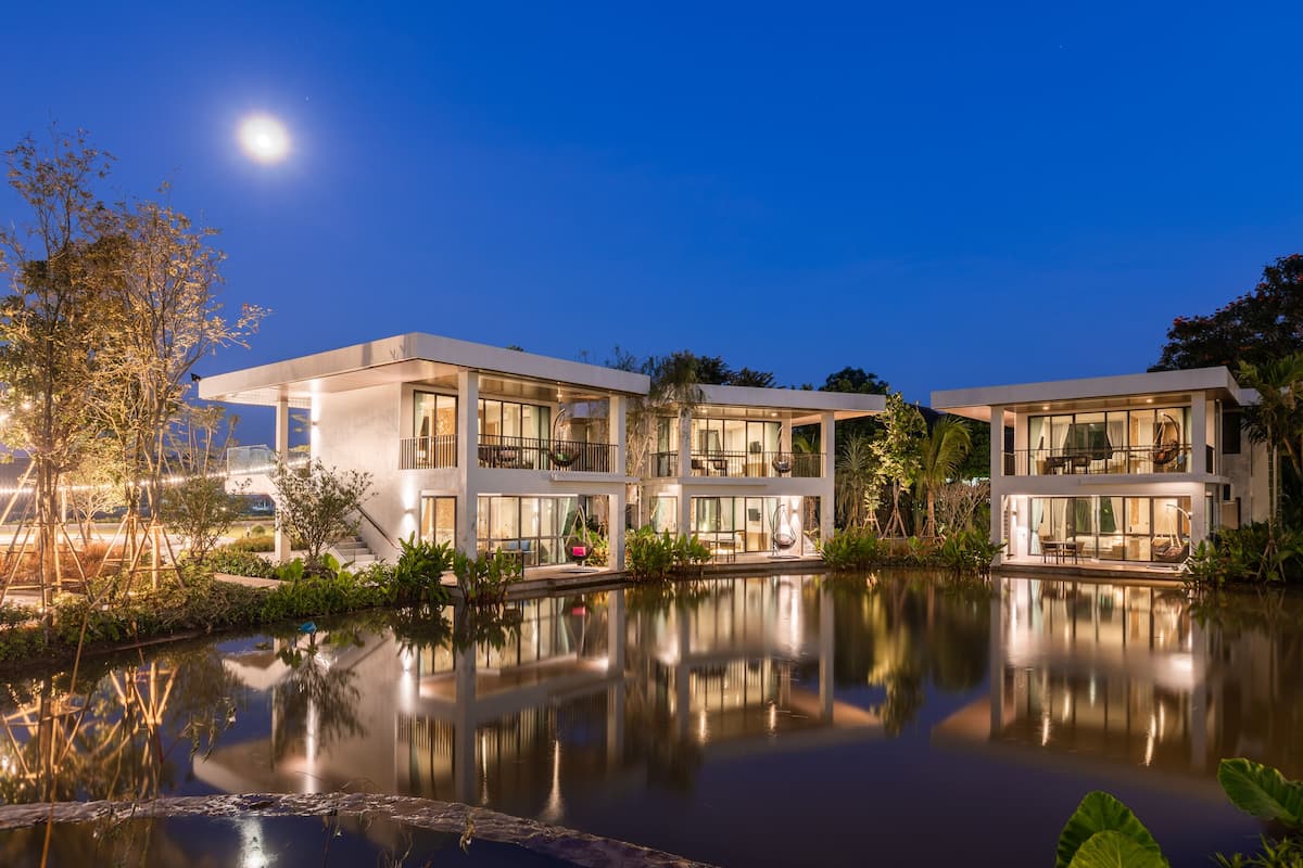 Um edifício moderno, de vários andares, com amplas janelas de vidro, um reflexo na água e a lua no céu.