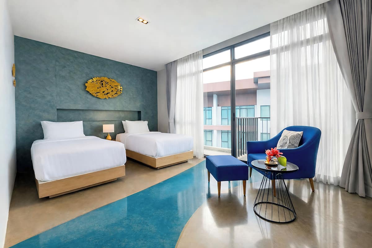 Quarto de hotel moderno com duas camas, uma poltrona azul, uma mesinha e vista para a cidade.