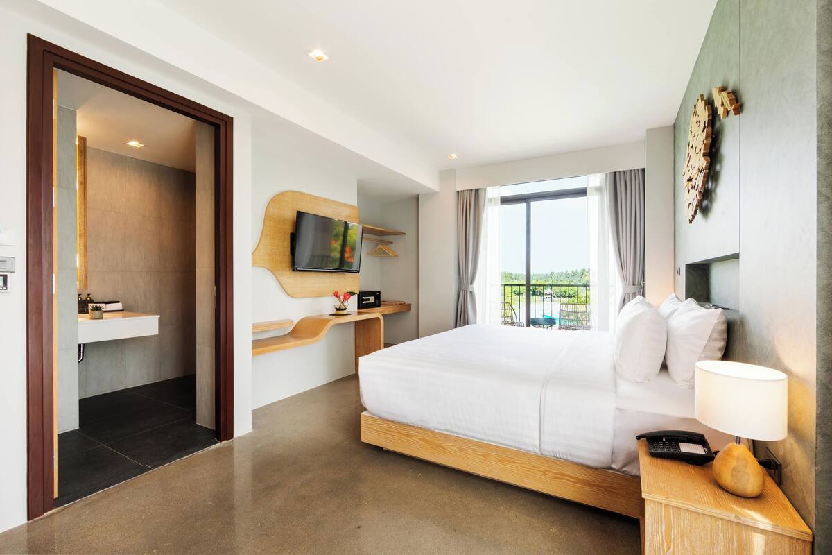 Quarto de hotel moderno com uma cama grande, mesa de cabeceira com abajur, televisão na parede e vista para o exterior.