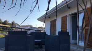 Uma área de estar coberta ao ar livre com móveis de vime escuro, um deck de madeira e um edifício com fachada branca e venezianas de madeira.