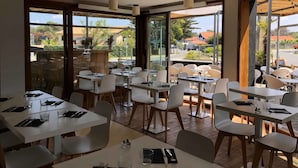 Um restaurante com mesas e cadeiras brancas, janelas amplas e vista para o exterior.