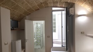 Quarto compacto com teto inclinado, pequena cozinha, cama e área de chuveiro com azulejos estampados.