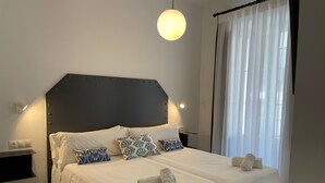 Uma cama de casal com cabeceira escura, roupa de cama branca, almofadas estampadas e mesa de cabeceira com abajur.