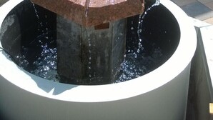 Um elemento decorativo com água, com topo de pedra e bacia circular, rodeado por um pátio pavimentado com bancos e mesas.