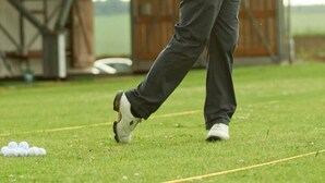 Uma pessoa jogando golfe em um campo gramado, com uma estrutura de madeira ao fundo.